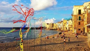 Uma praia com pipas coloridas, um edifício de pedra e o mar.