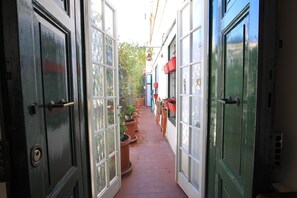 Um corredor estreito com portas abertas que dão para uma área de jardim, plantas em vasos e um toldo vermelho.