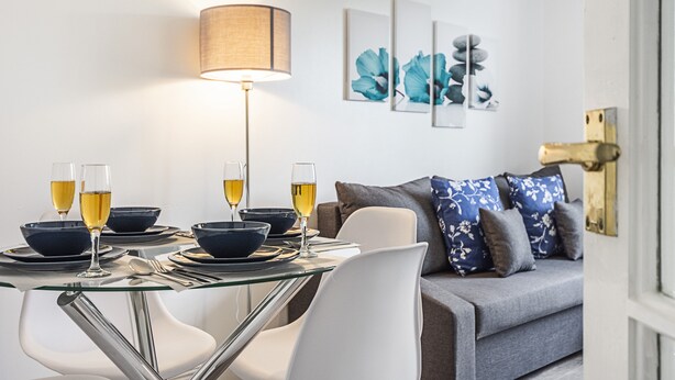 Uma sala de jantar moderna com uma mesa de vidro, cadeiras brancas e uma luminária. Há três tigelas e copos com uma bebida amarela sobre a mesa.