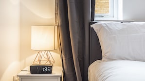 Uma cama bem arrumada com roupa de cama branca, uma mesa de cabeceira com um relógio digital e uma luminária.