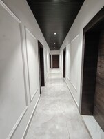 Um corredor comprido e estreito, com paredes brancas e chão de mármore, que termina numa porta de madeira escura.