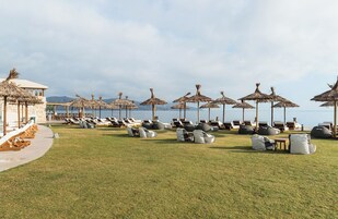 Uma área de estar à beira-mar com espreguiçadeiras e guarda-sóis de palha, um edifício de pedra e vista para o mar.