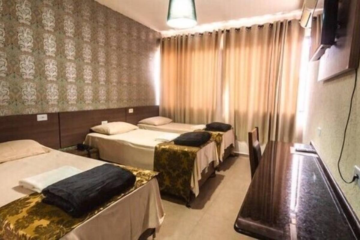 Quarto de hotel com duas camas de solteiro, cômoda de madeira, televisão e janela com cortinas.