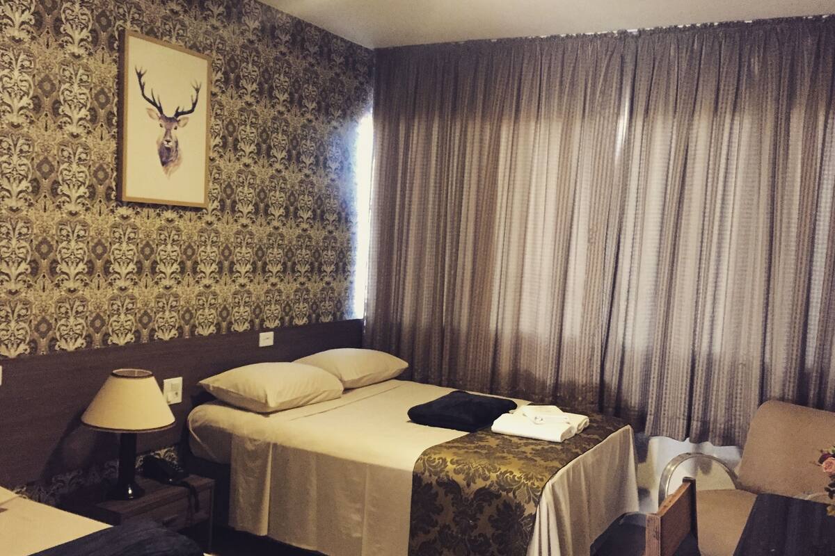 Quarto de hotel com papel de parede estampado, uma cama com cabeceira, mesa de cabeceira com abajur, uma cadeira e uma janela grande com cortinas.