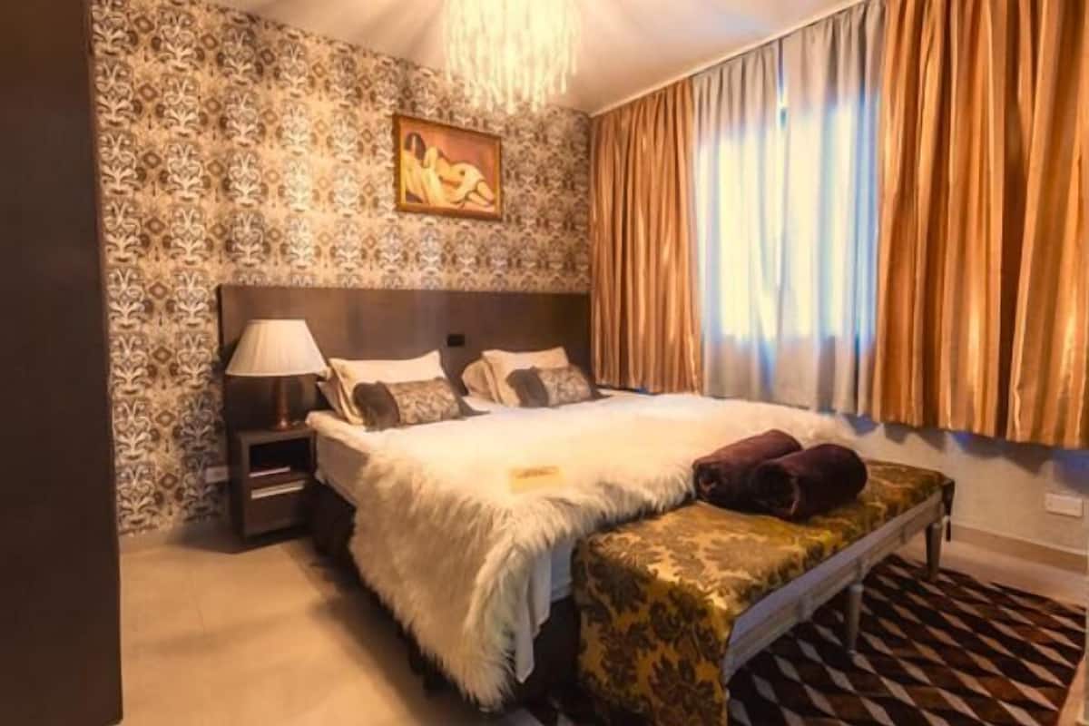 Quarto com papel de parede estampado, uma cama com edredom branco, uma mesinha de cabeceira, um abajur e um banco com capa estampada.