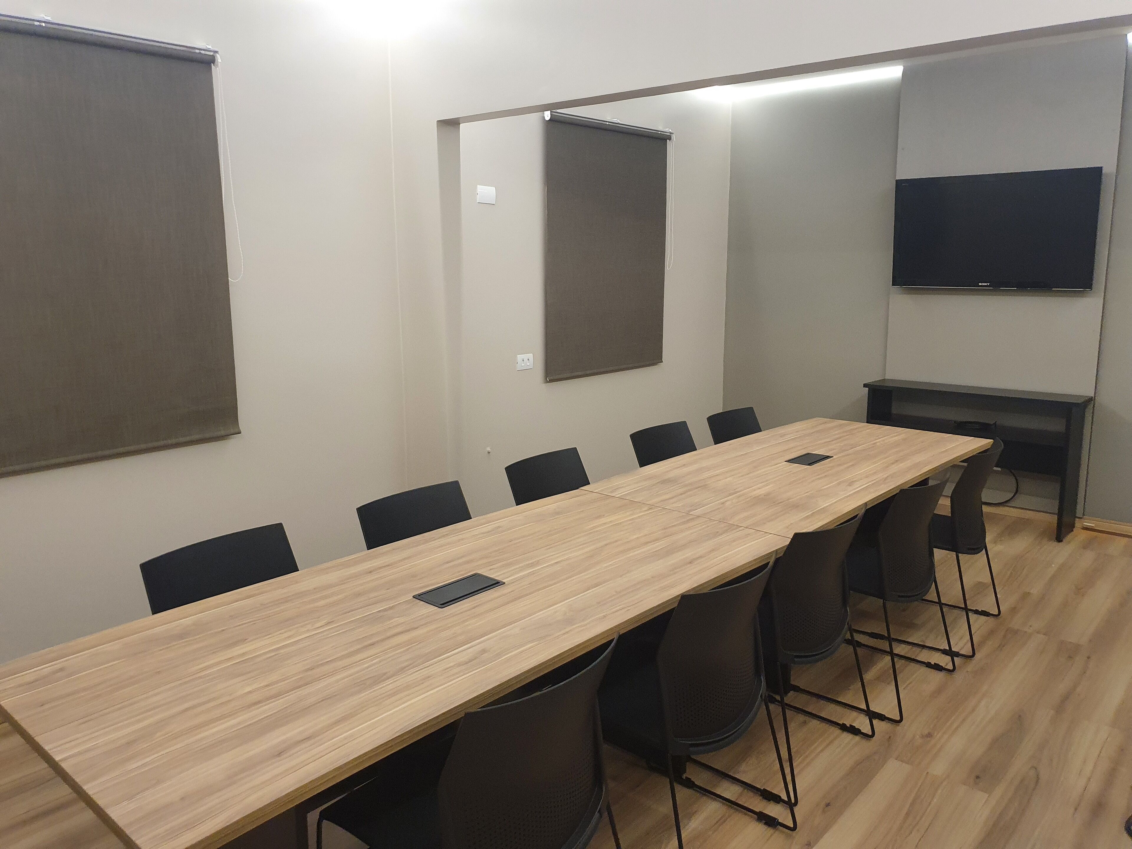 Uma sala de conferência com uma mesa de madeira comprida, cadeiras pretas, uma tela de projetor e uma TV de tela plana na parede.