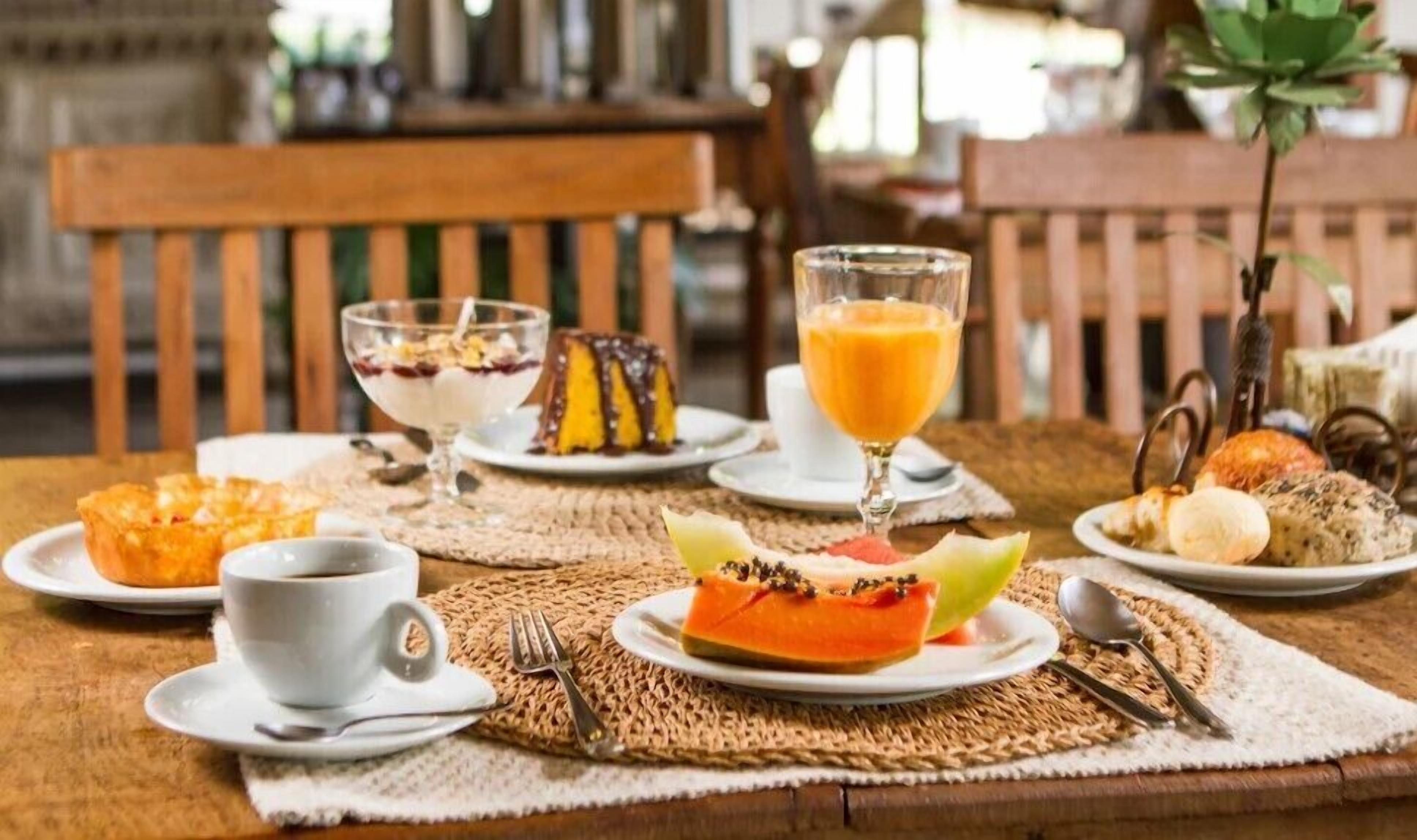 Disposição para o pequeno-almoço com vários pratos, um copo de sumo de laranja e uma chávena de café sobre uma mesa de madeira.