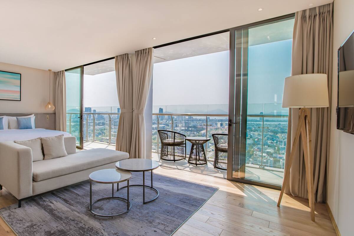 Quarto de hotel moderno com uma cama grande, um sofá, duas mesinhas e uma varanda com vista para a cidade.