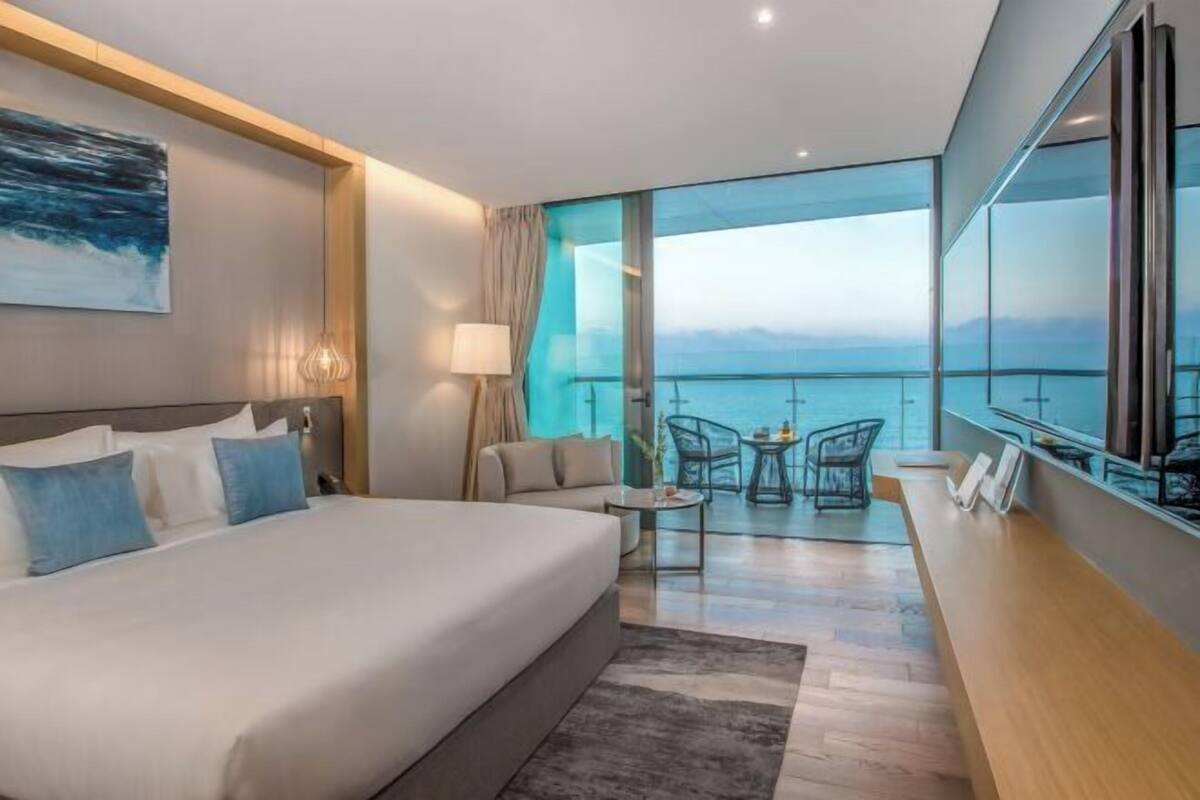 Quarto de hotel moderno com uma cama grande, área de estar com vista para o mar e varanda com paredes de vidro.