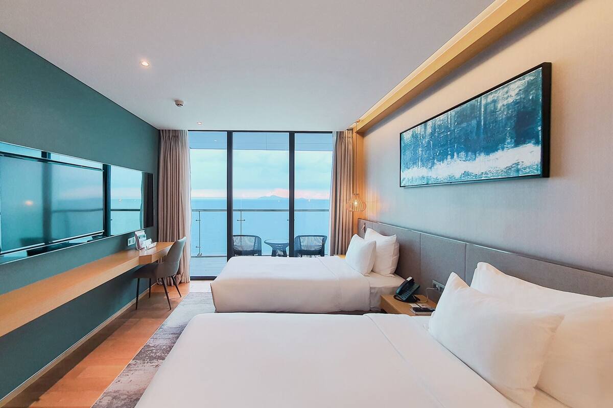 Quarto de hotel com duas camas, uma escrivaninha com cadeira, televisão e uma janela grande com vista para o mar.
