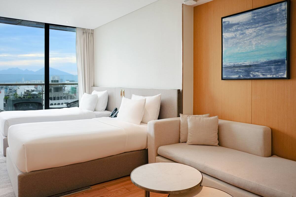 Quarto de hotel com duas camas, um sofá, uma mesa redonda e uma janela grande com vista para a cidade.