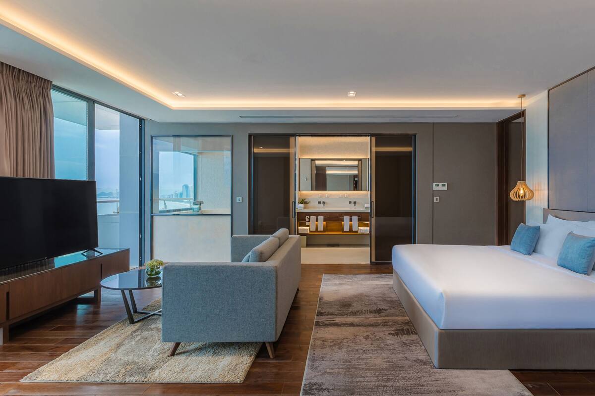 Quarto de hotel moderno com uma cama grande, área de estar, televisão e banheiro com toucador.