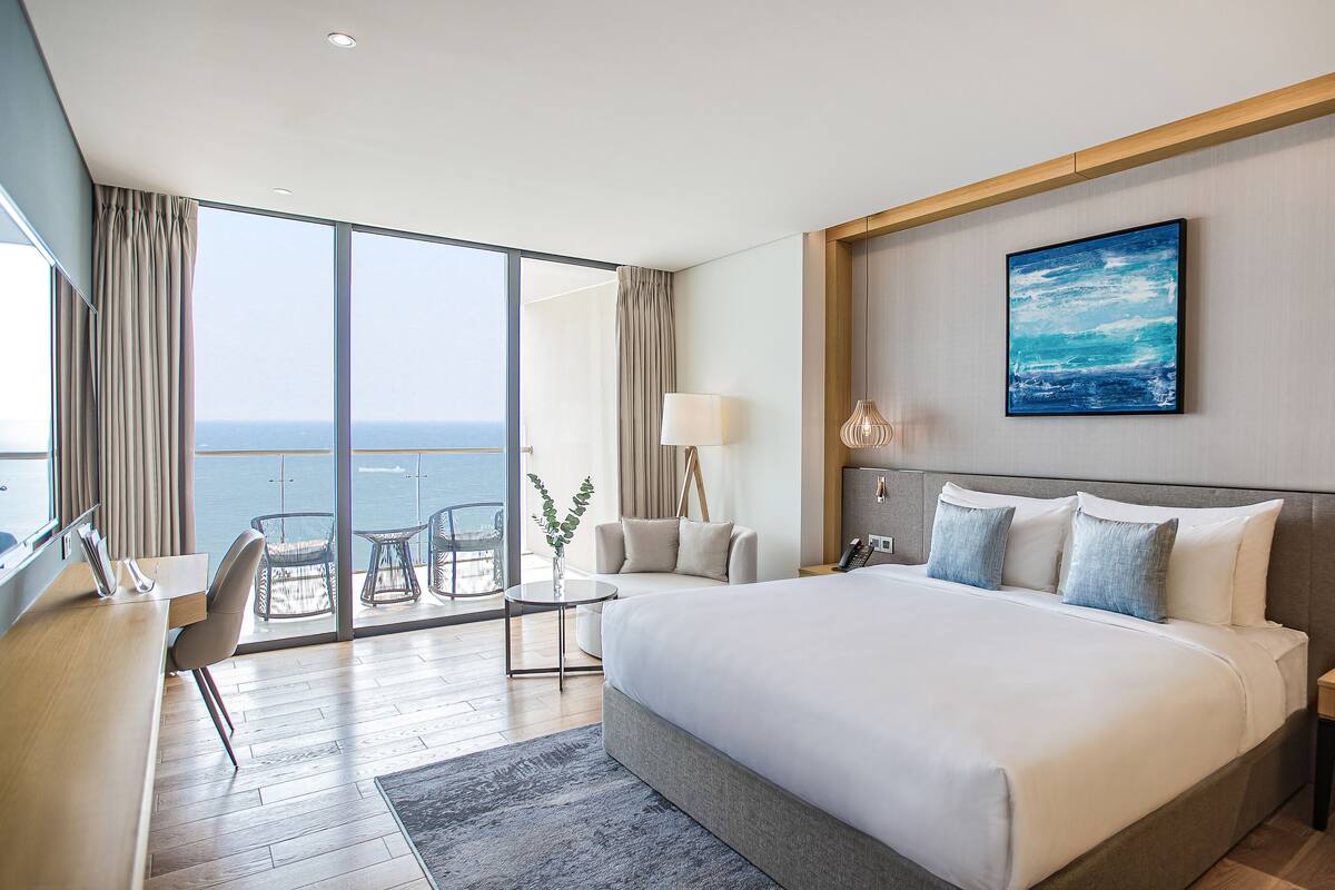 Quarto de hotel moderno com uma cama grande, uma escrivaninha e uma varanda com vista para o oceano.