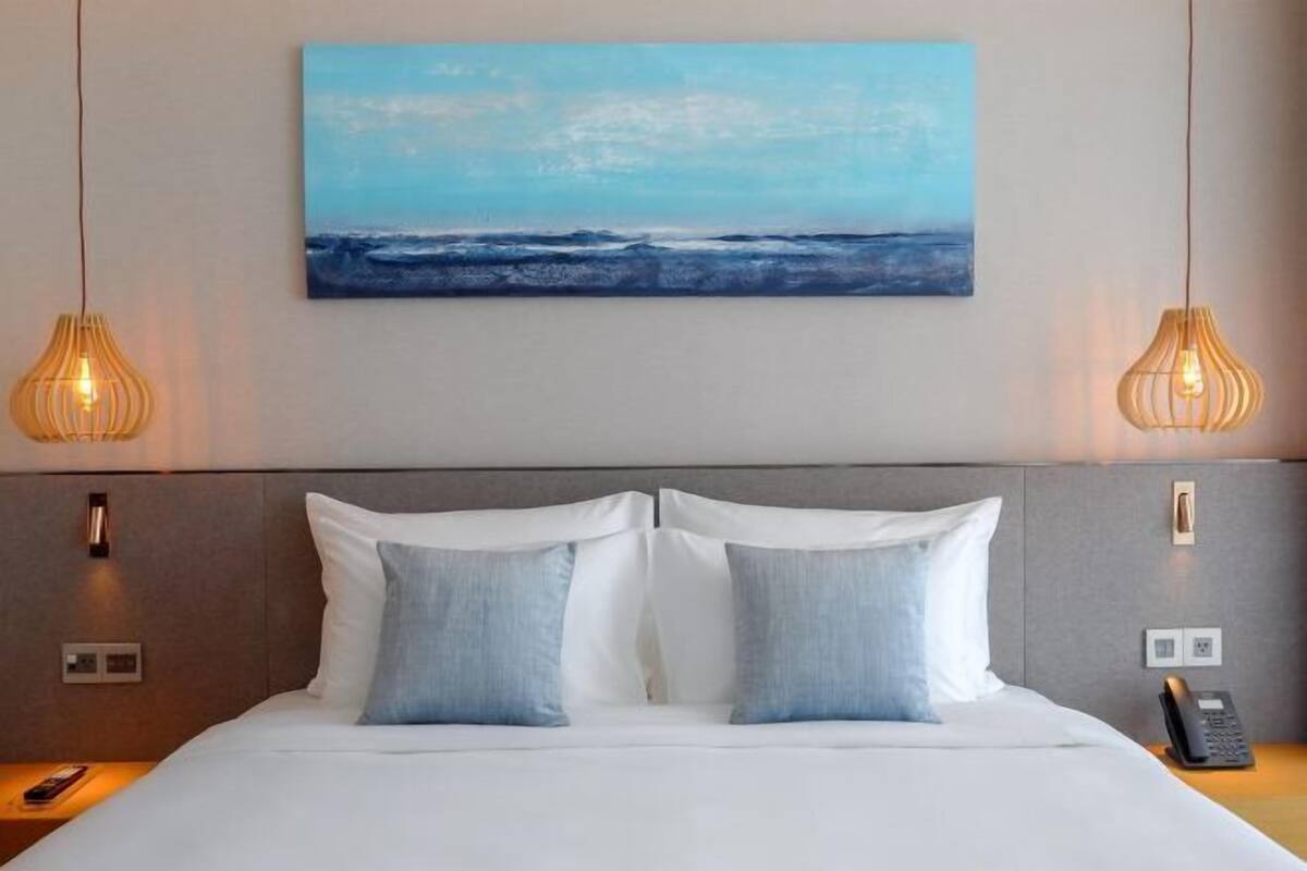 Quarto de hotel com uma cama, duas luminárias de cabeceira, um quadro com vista para o oceano e um telefone no criado-mudo.