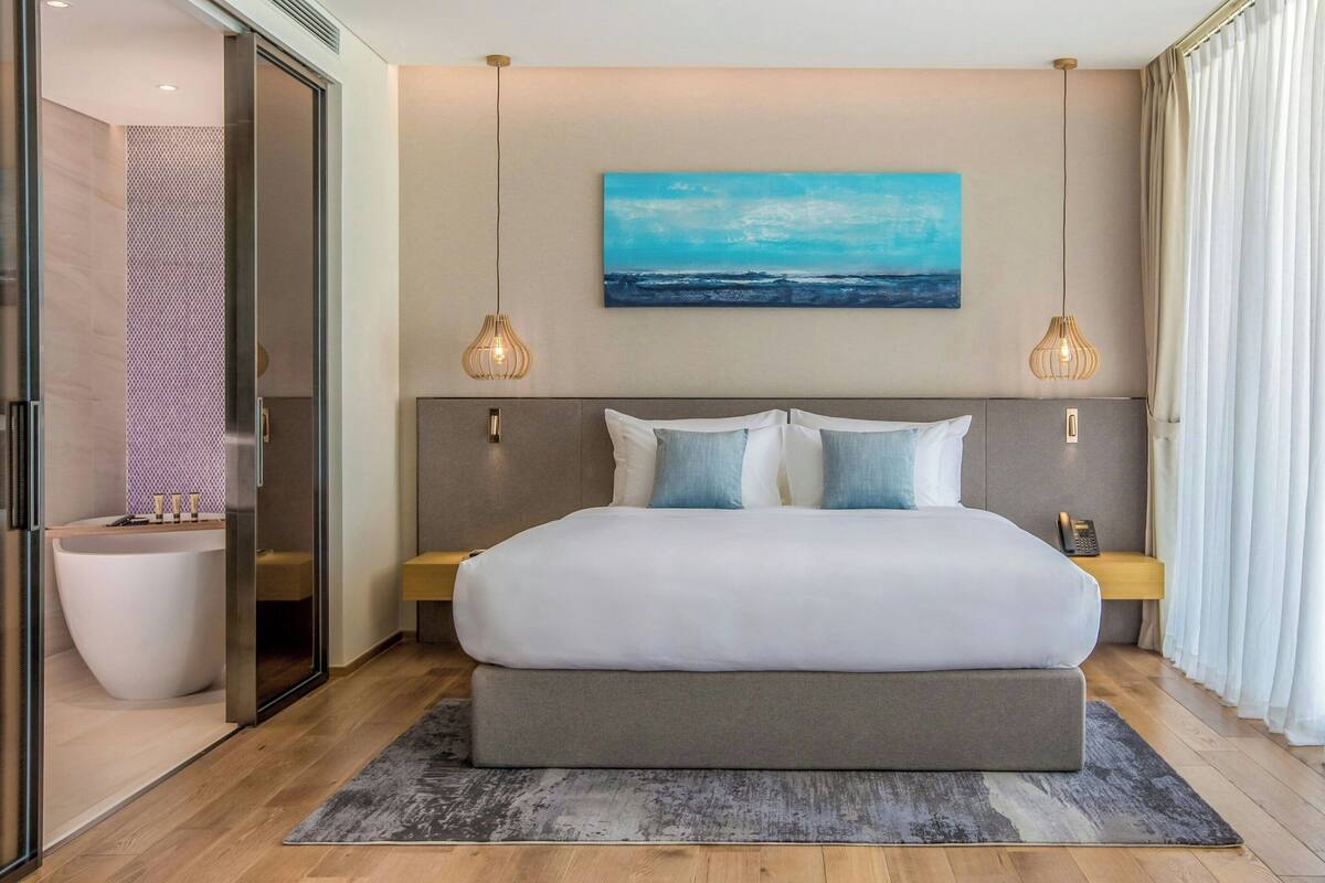 Quarto de hotel moderno com uma cama grande, duas mesas de cabeceira, um quadro sobre a cama e vista para o mar pela janela.