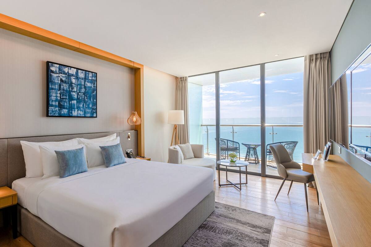 Quarto de hotel moderno com uma cama grande, uma escrivaninha, uma cadeira e uma varanda com vista para o mar.