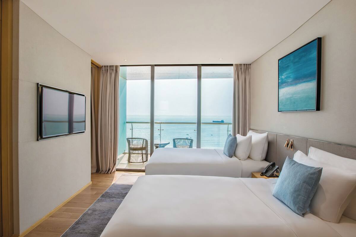 Quarto de hotel com duas camas, uma televisão, um quadro e varanda com vista para o mar.