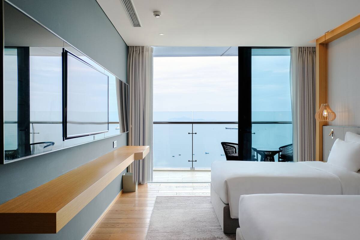 Quarto de hotel moderno com uma cama grande, televisão na parede, varanda com vista para o mar e um banco de madeira.