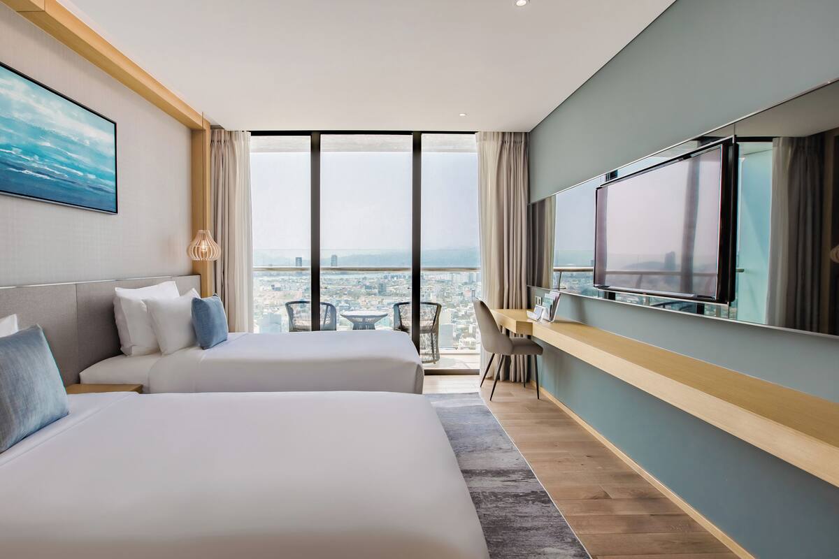 Quarto de hotel com duas camas, uma televisão grande e varanda com vista para a cidade.