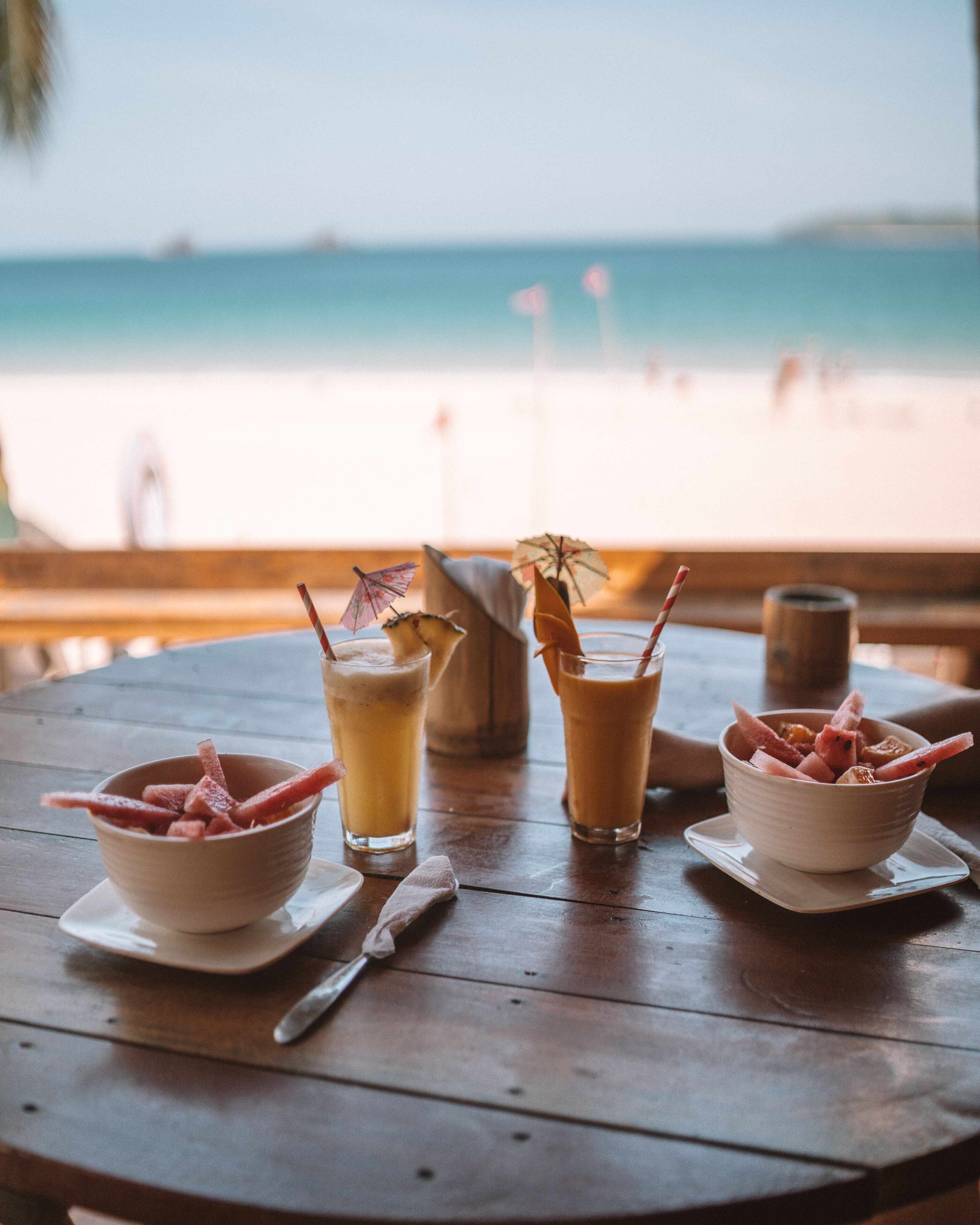 Uma mesa de madeira com uma tigela de frutas, dois copos de uma bebida amarela e uma jarra, com vista para a praia.
