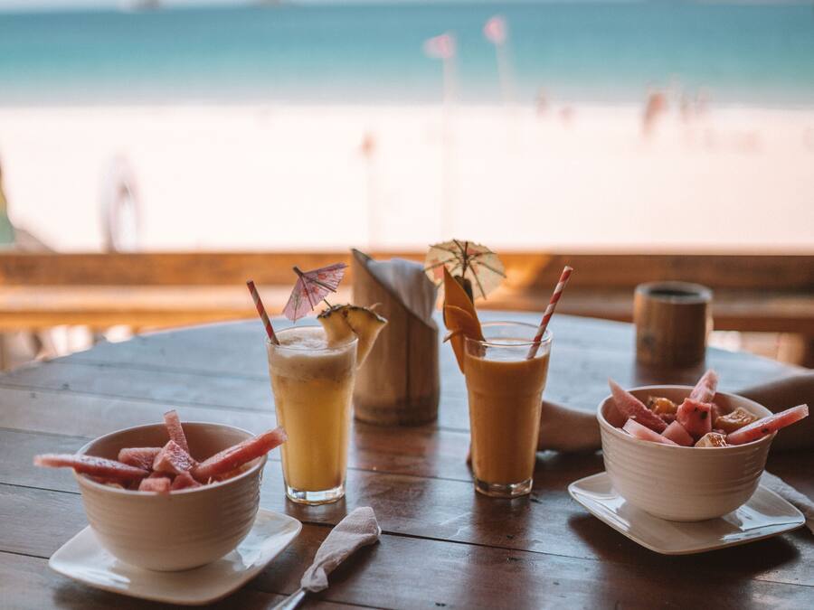 Uma mesa de madeira com uma tigela de frutas, dois copos de uma bebida amarela e uma jarra, com vista para a praia.