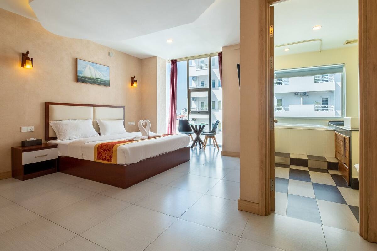 Quarto de hotel moderno com uma cama grande, mesas de cabeceira, uma escrivaninha e vista da cidade através de amplas janelas.