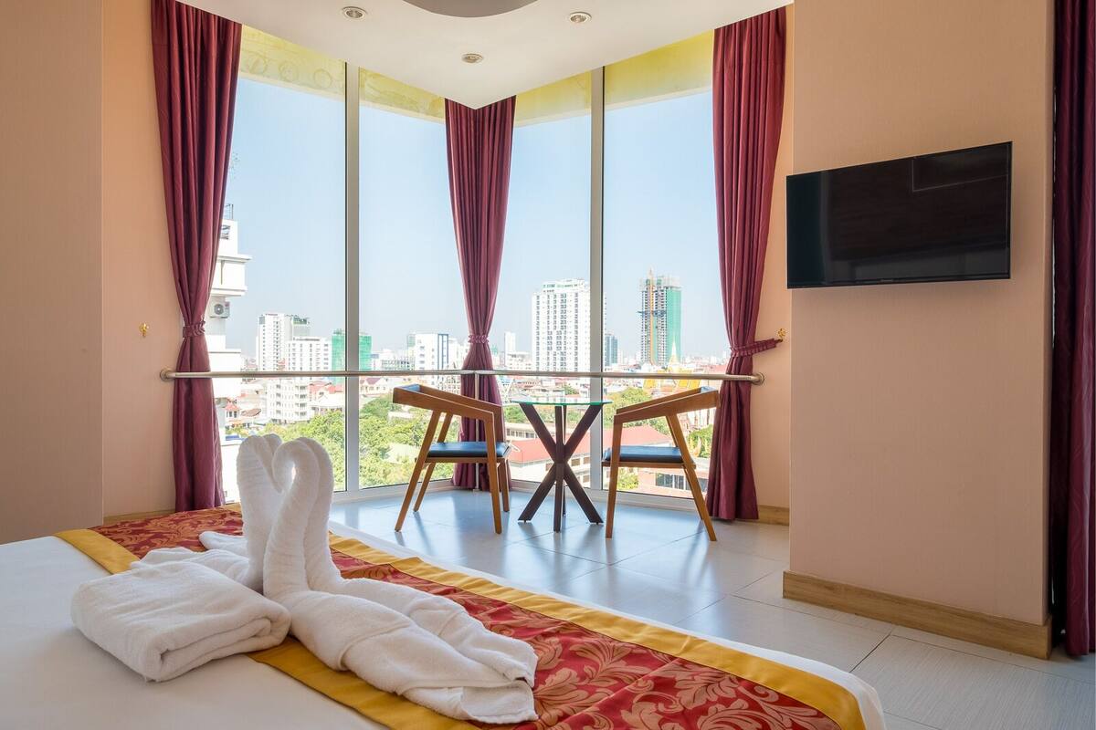 Quarto de hotel com cama, mesa e cadeiras, televisão e varanda com vista para a cidade.