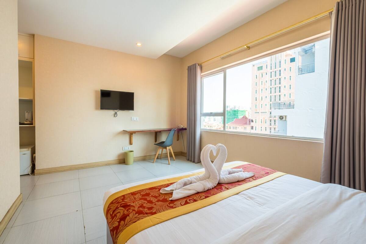 Quarto de hotel com cama, escrivaninha, cadeira, televisão e janela com vista para edifícios.