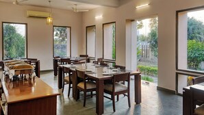 Uma sala de jantar com uma longa mesa de madeira posta para uma refeição, rodeada por cadeiras. Há janelas amplas que oferecem vista para o verde lá fora.