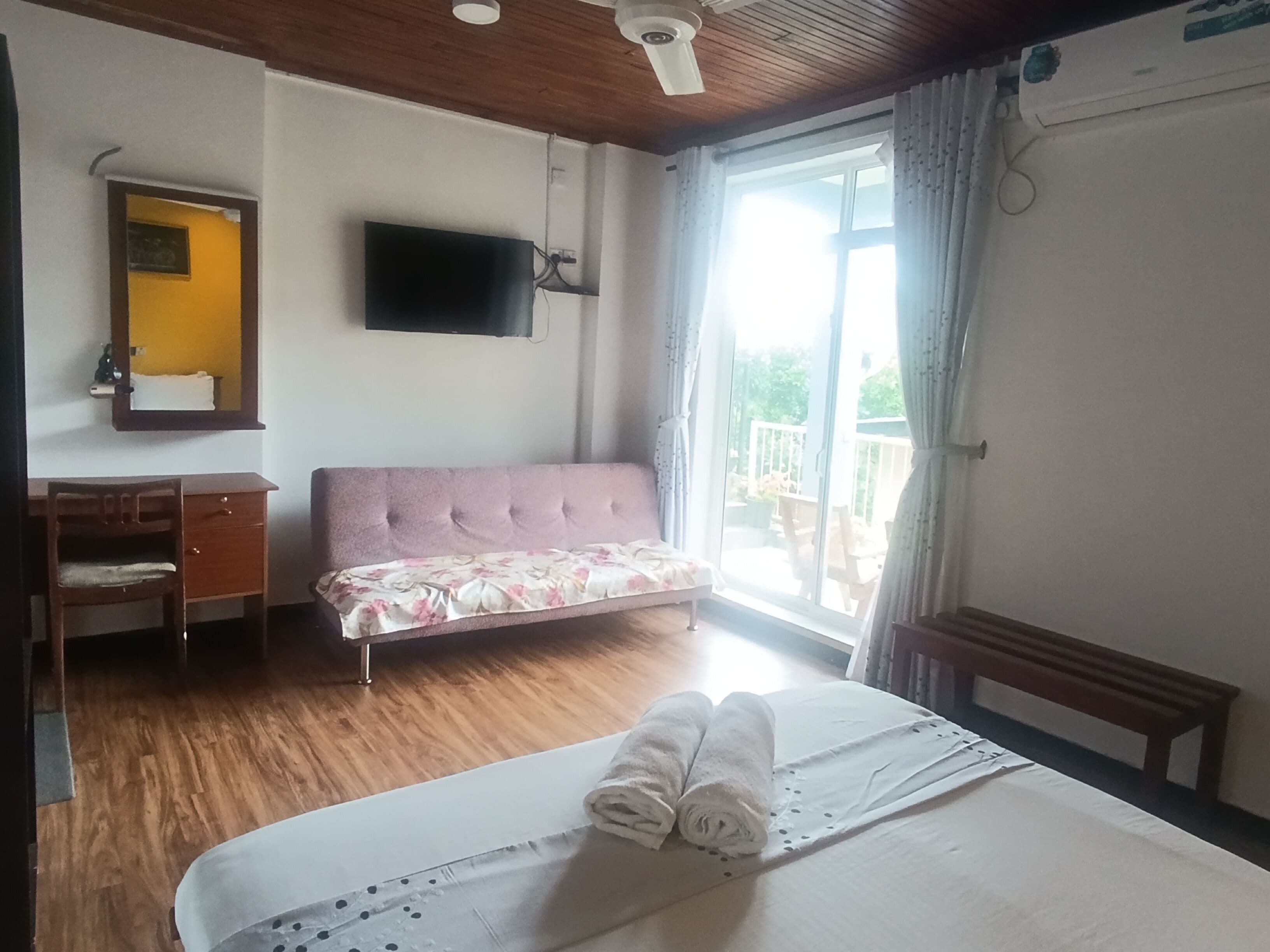 Quarto com cama, escrivaninha, cadeira, televisão e varanda.
