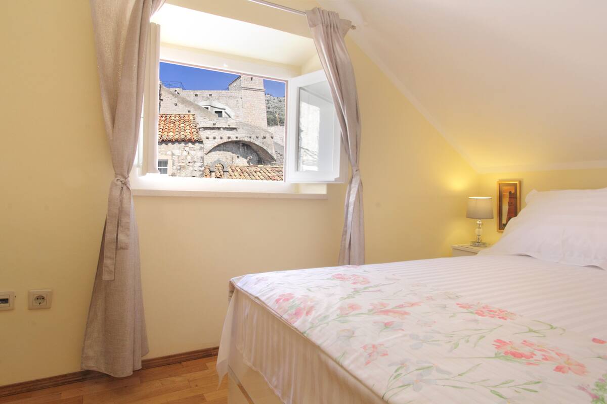 Quarto com cama, janela com cortinas, mesa de cabeceira com abajur e vista para uma ponte de pedra.