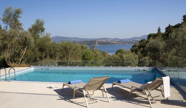 Uma piscina com águas azuis cristalinas, rodeada por espreguiçadeiras e vista para o mar e as colinas.