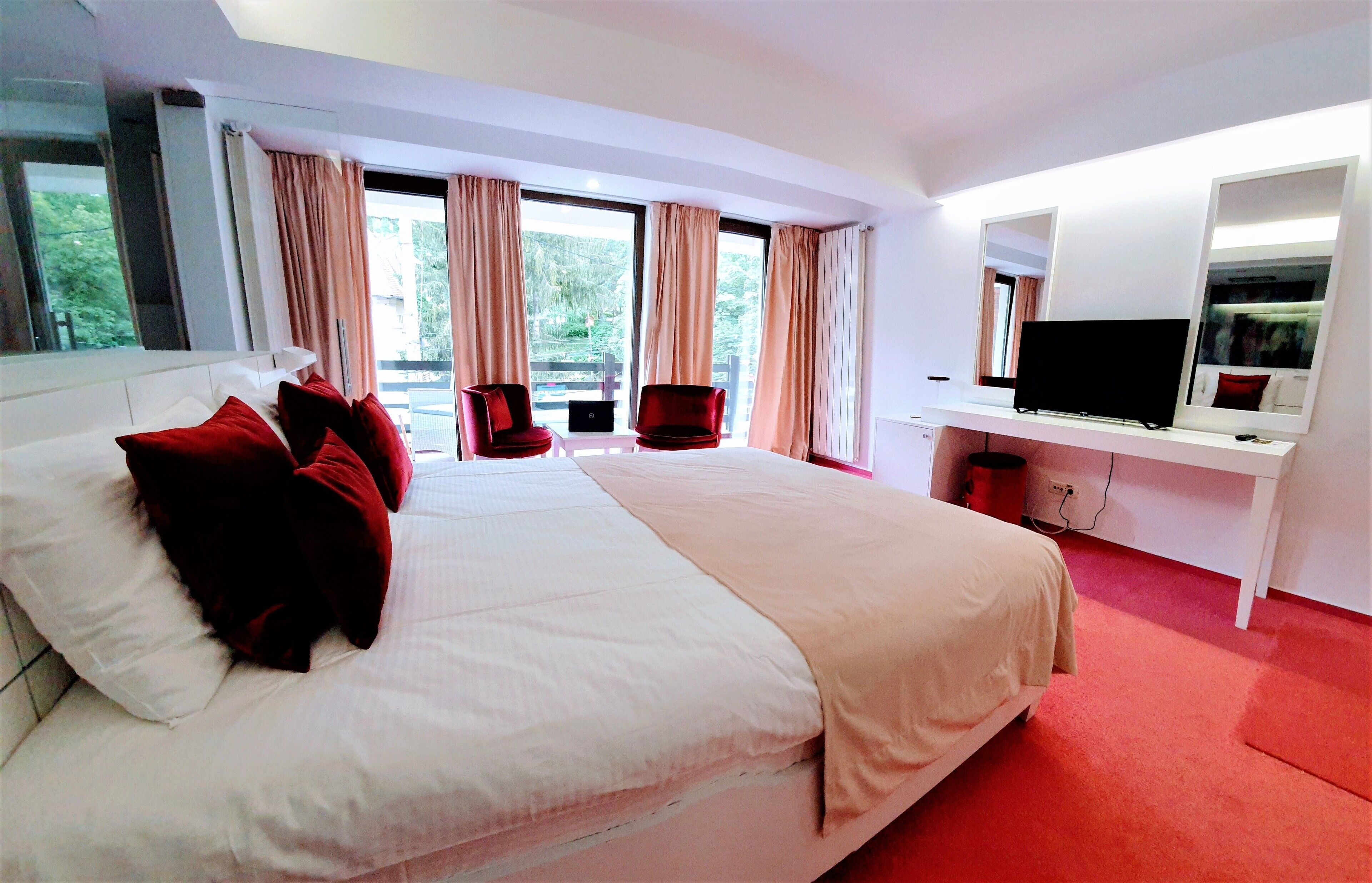 Quarto de hotel moderno com uma cama grande, cadeiras vermelhas, uma televisão e uma varanda com cortinas.