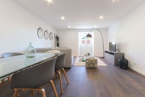 Uma sala de estar moderna com uma mesa de jantar de vidro, sofás cinzas, um banquinho estampado e uma TV em um móvel.
