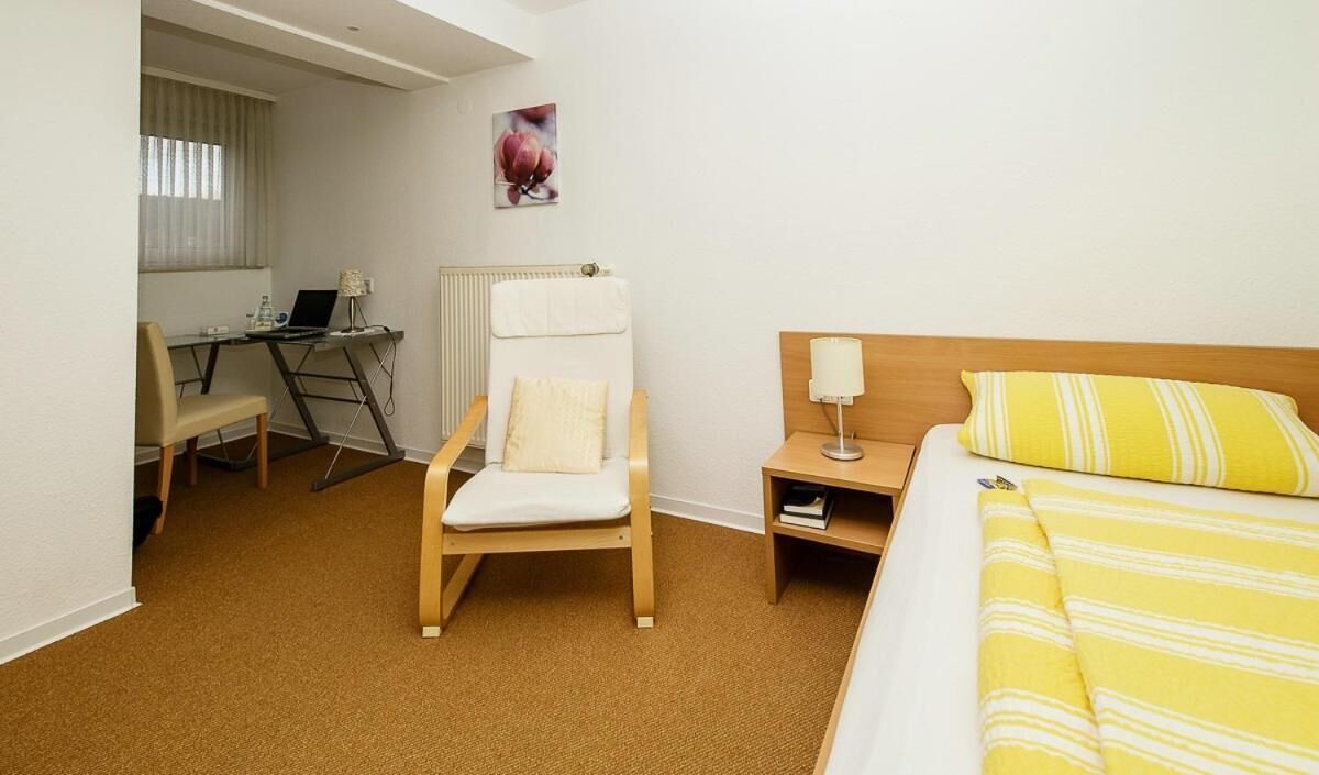 Quarto de hotel com cama, uma cadeira, escrivaninha com um laptop, radiador e um quadro na parede.