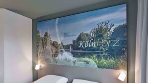 Quarto de hotel com uma janela grande mostrando uma vista panorâmica de um lago e árvores, e a inscrição 'Köln Porz' na janela.