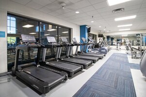 Uma academia com várias esteiras alinhadas em uma parede, piso coberto por um tapete azul e diversos equipamentos de exercício ao fundo.