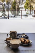 Mesa com xícara de café, caneca e um doce, numa varanda com vista para uma paisagem nevada.