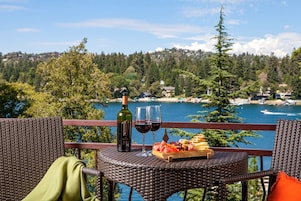 Uma mesa de vime com uma garrafa de vinho, copos e uma tábua de frios, com vista para um lago e casas ao fundo.