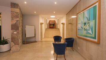 Um hall de hotel com parede de destaque em pedra, uma escadaria e dois poltronas azuis.