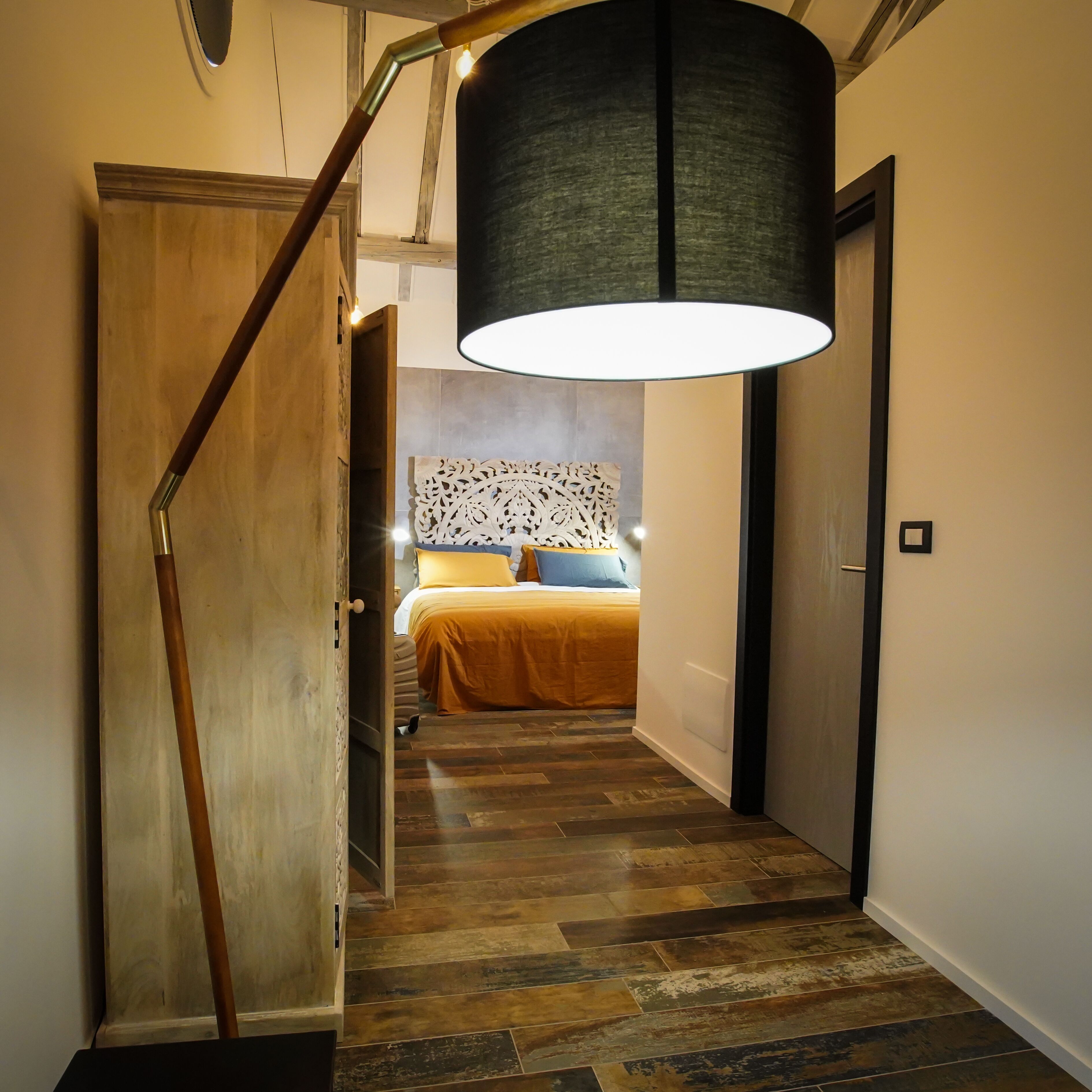 Um corredor com escada de madeira, cama com cabeceira estampada e um abajur grande.