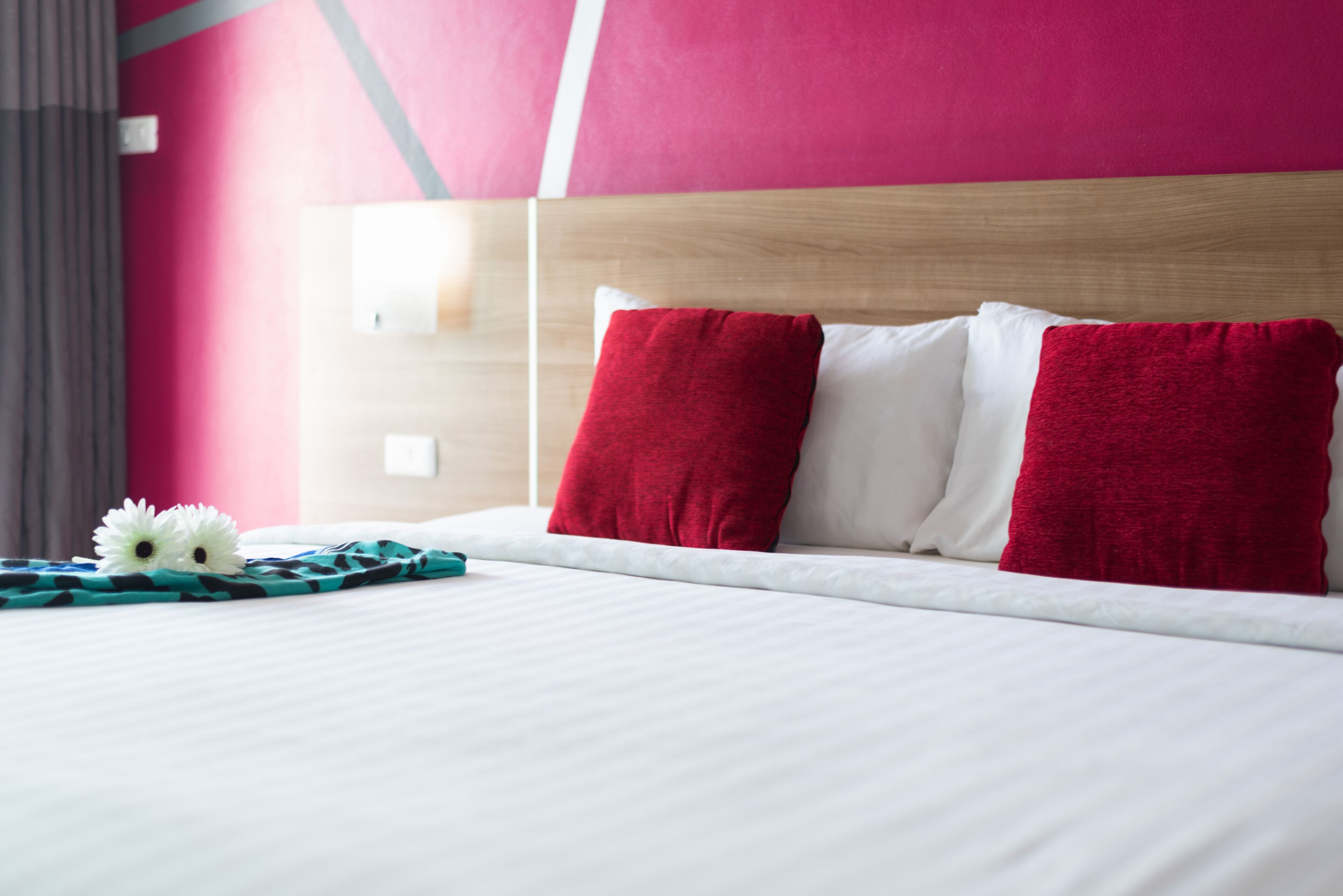 Quarto de hotel com cama, almofadas vermelhas e brancas, manta branca e azul-turquesa e uma flor branca.