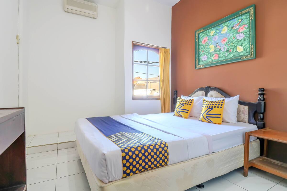 Cama bem arrumada com roupa de cama estampada, janela com cortinas e um quadro pendurado na parede.