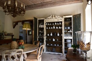 Uma área de jantar rústica com uma mesa de madeira, cadeiras e um candelabro. Há prateleiras com louças e um vaso com rolhas.