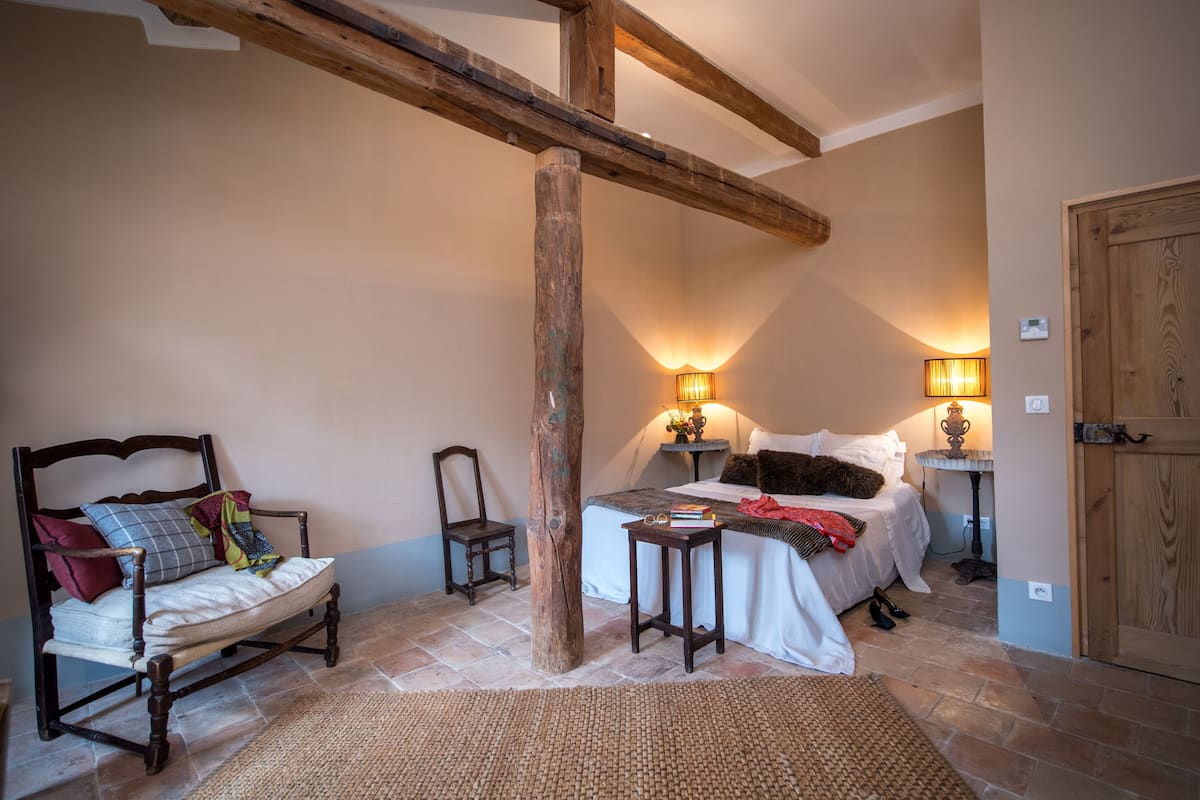Quarto com cama, uma cadeira, um poste de madeira e duas luminárias fixadas na parede.
