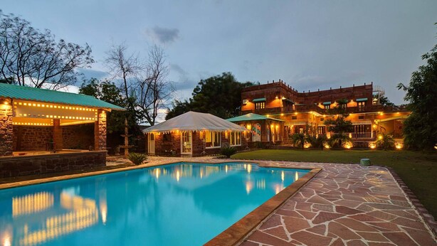 Uma piscina com ladrilhos de pedra, um edifício de pedra com telhado branco e uma grande casa de tijolos com várias janelas e luzes.