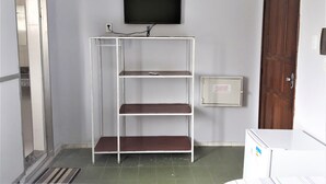 Um quarto com uma estante de metal branca, uma televisão fixada na parede, uma porta de madeira e uma pequena caixa elétrica.