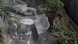 Um mural retrata uma árvore e duas figuras em um barco, com móveis de jardim e plantas em primeiro plano.