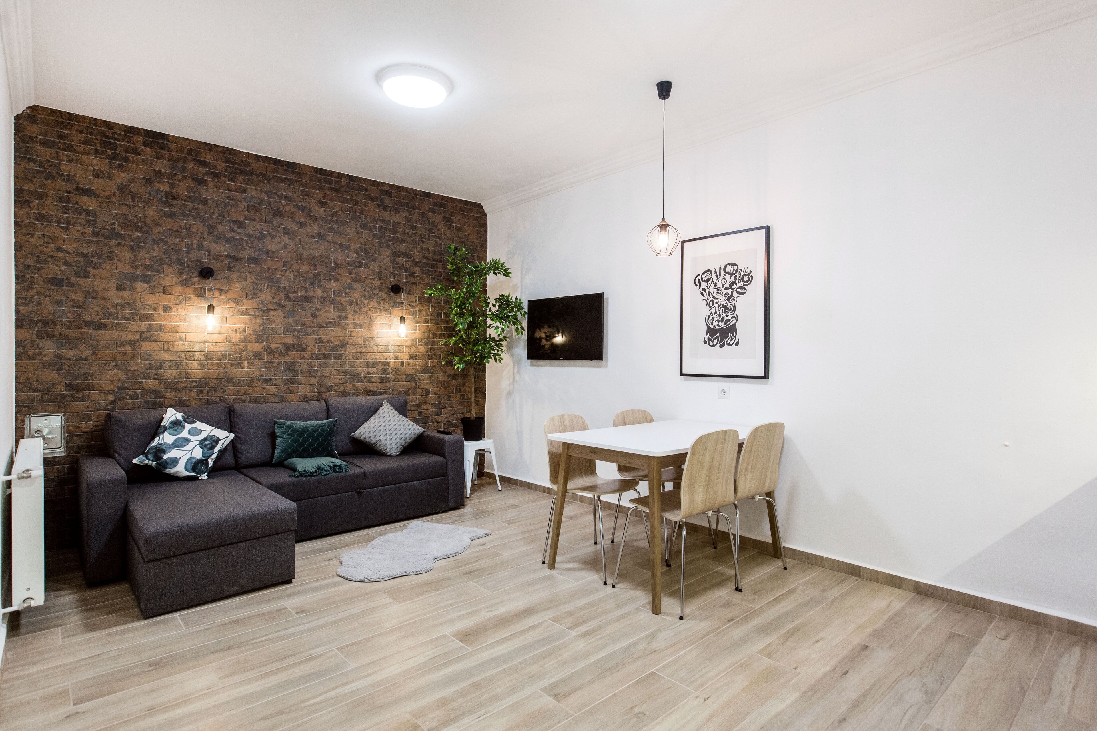 Uma sala de estar moderna com parede de tijolos decorativa, um sofá cinza com almofadas estampadas, uma mesa de jantar de madeira com cadeiras e uma TV de tela plana na parede.