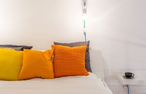 Cama com três almofadas em tons de amarelo, laranja e castanho, mesa de cabeceira branca com uma chávena preta, e uma tomada.
